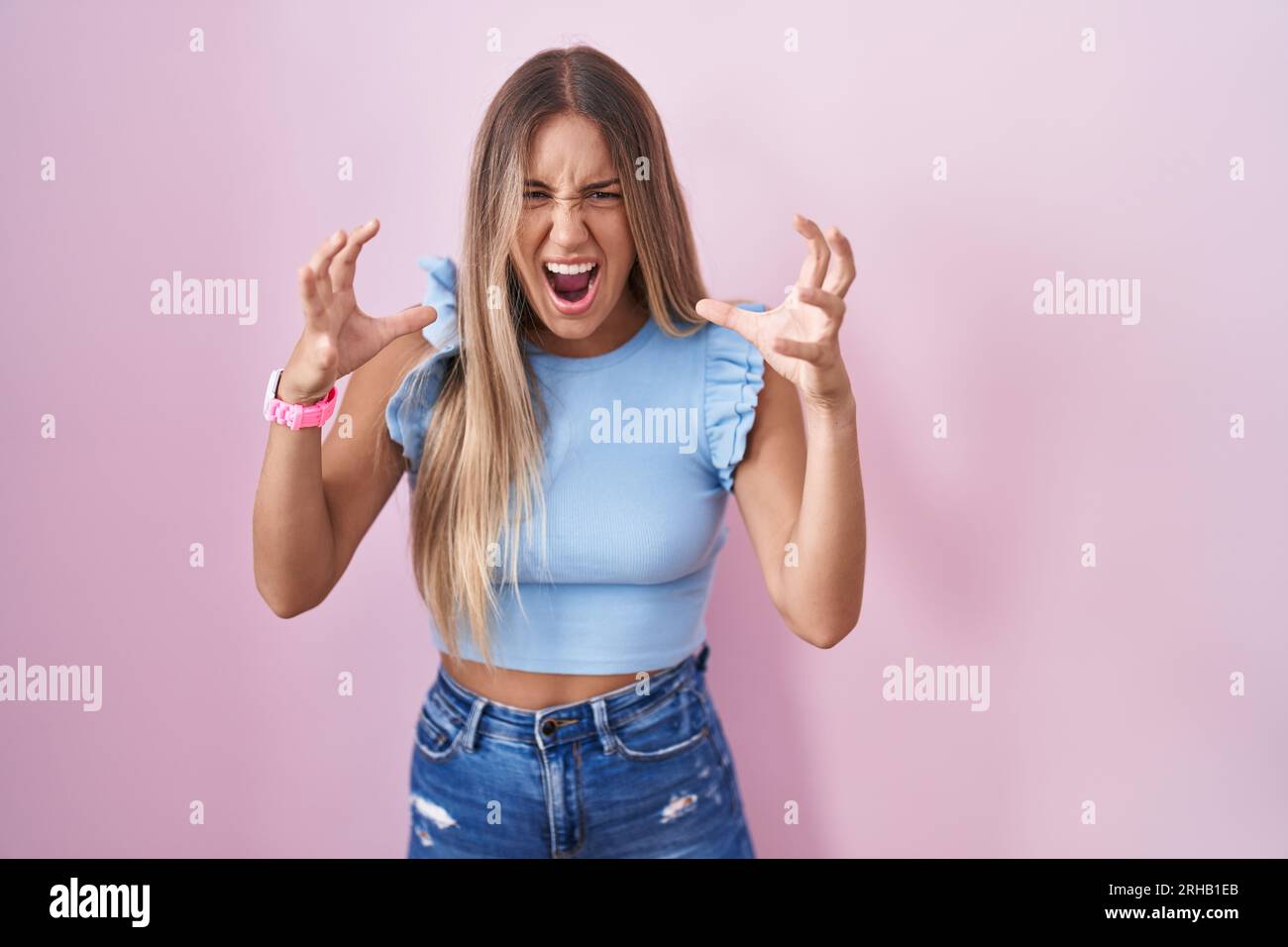 Young blonde woman standing over pink background shouting frustrated ...