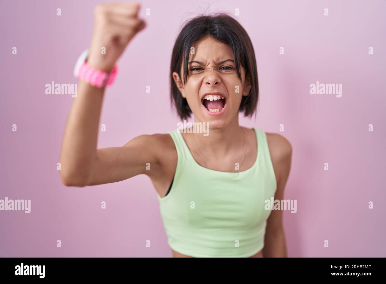 Young girl standing over pink background angry and mad raising fist ...