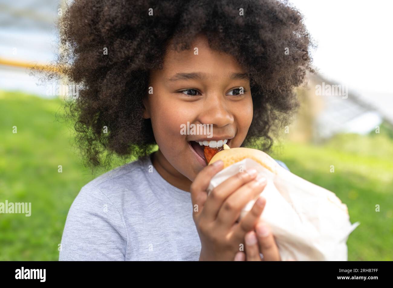 Dark skinned female child eating junk food with pleasure in park while ...