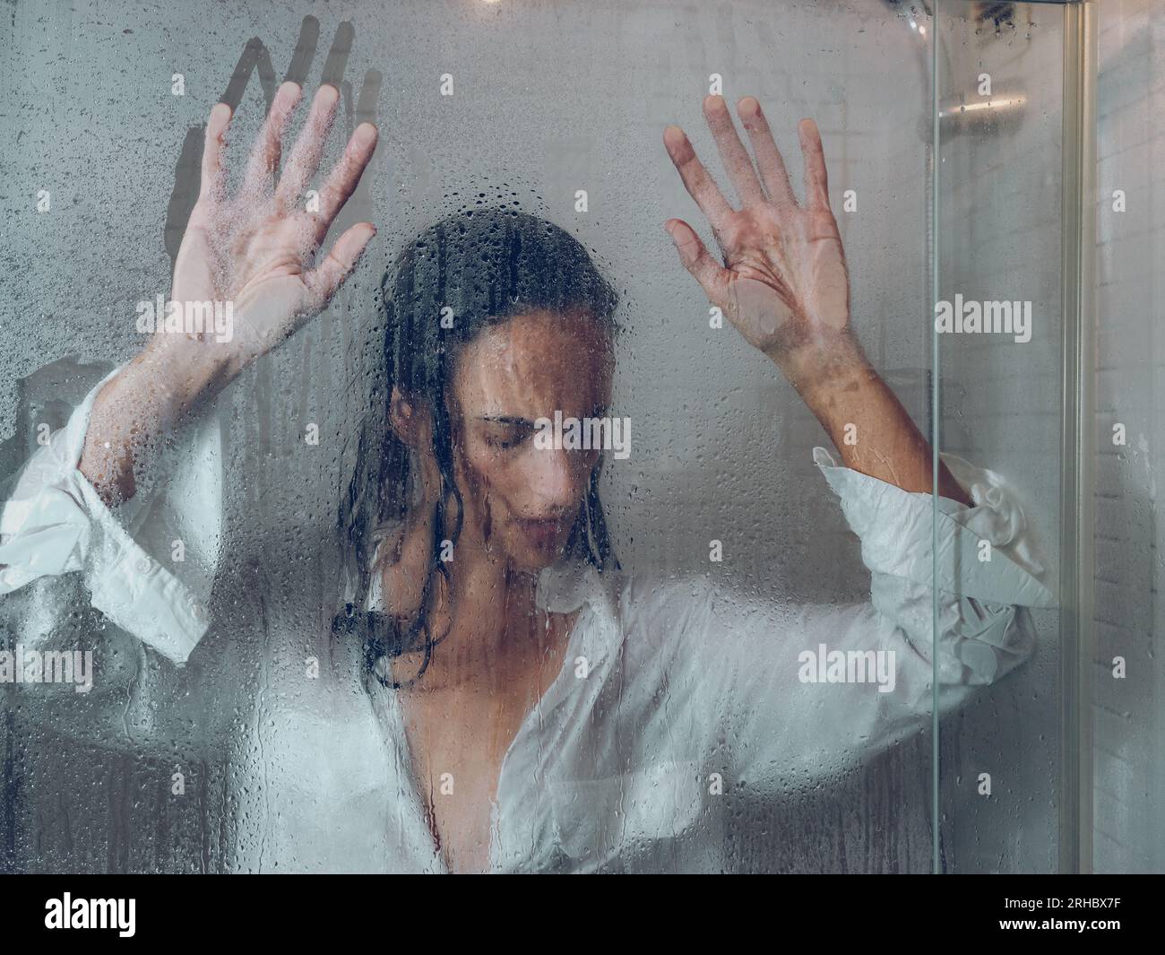 Through glass of sad female with black hair in wet wear touching ...