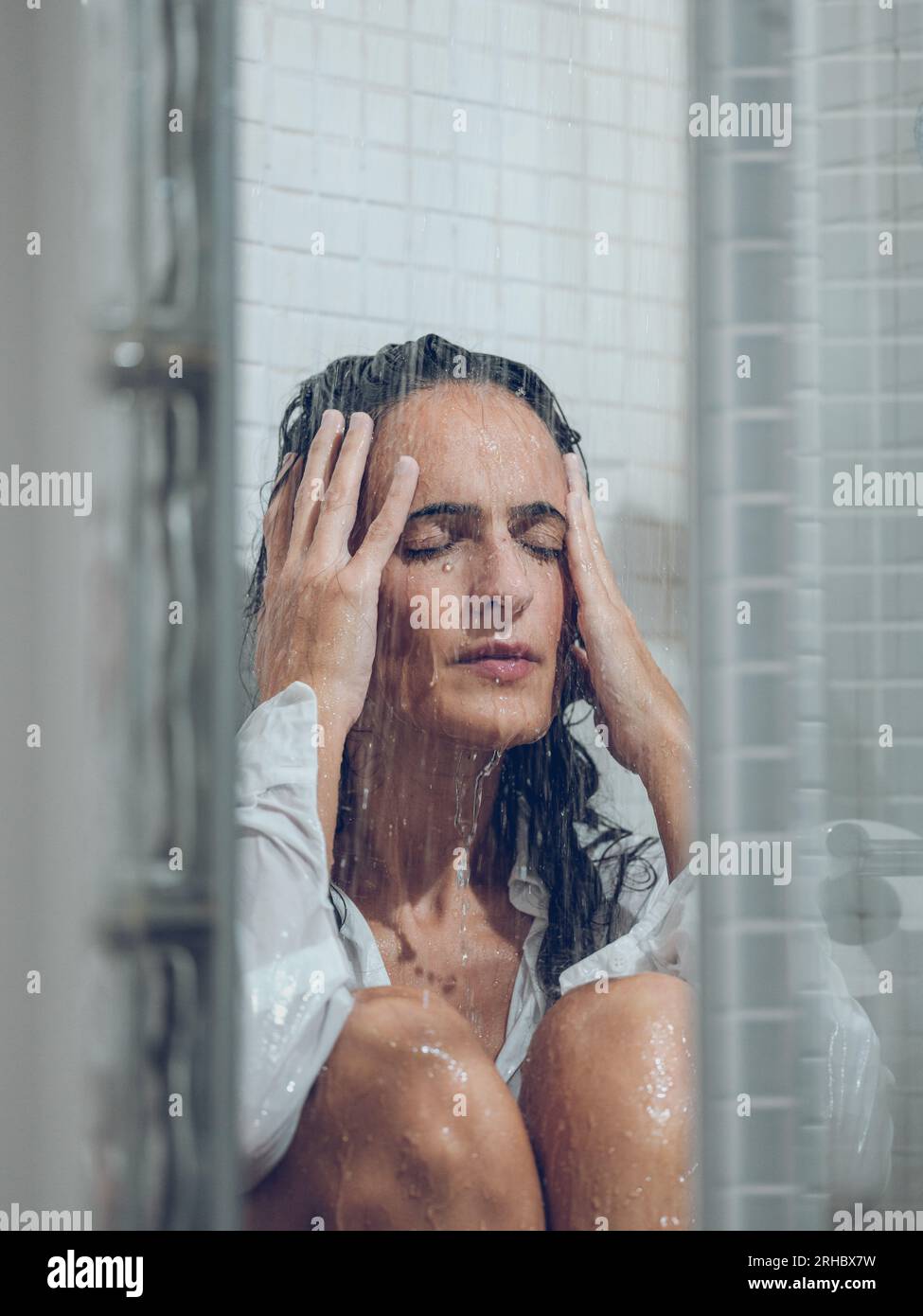 Depressed Woman With Dark Hair In Wet White Shirt And With Closed Eyes Sitting In Shower Under