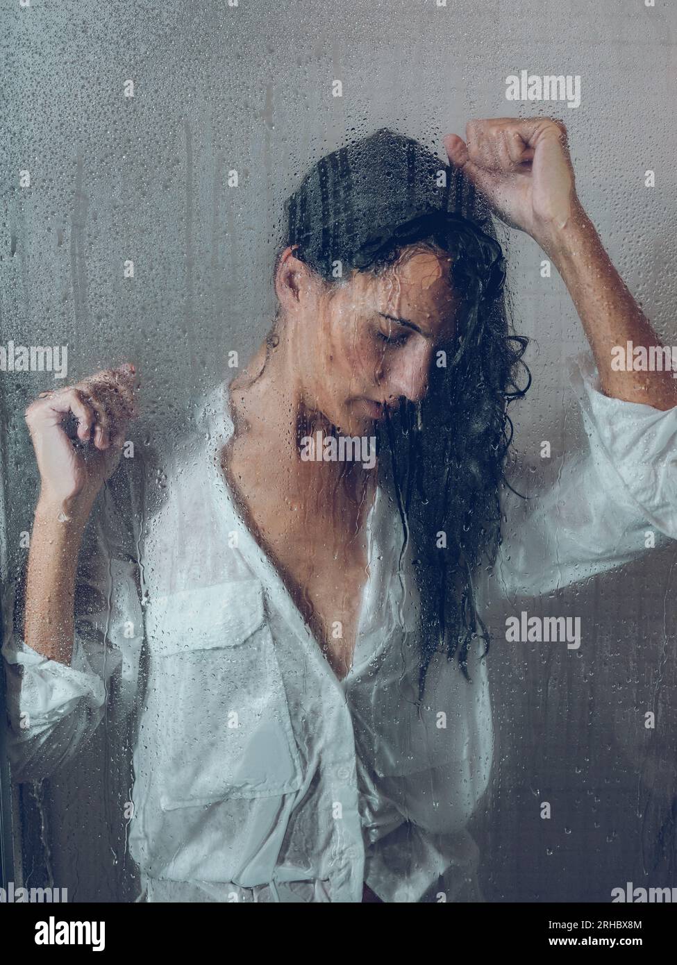 Through glass of sad female with black hair in white wet shirt standing ...