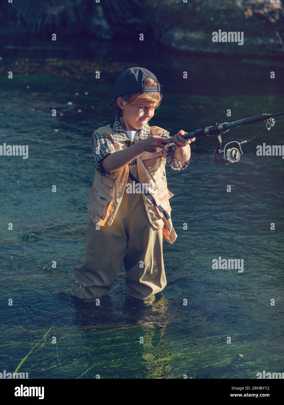 Concentrated boy in waterproof pants and cap pulling fish with rod ...