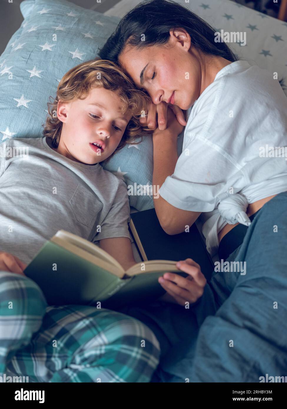 From above of adorable little boy reading interesting book lying on bed ...
