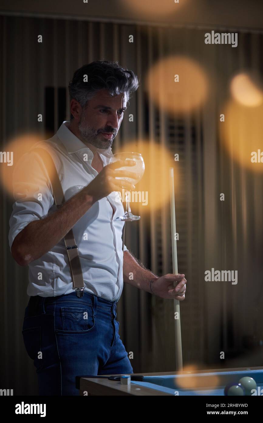 Serious bearded man with cue standing at pool table and drinking ...