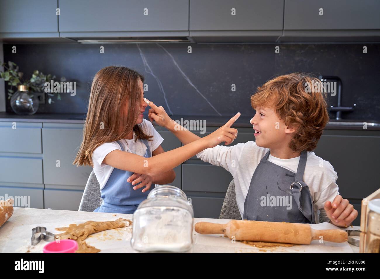 Cheerful brother and sister touching noses of each other with index fingers dirty of flour while ...