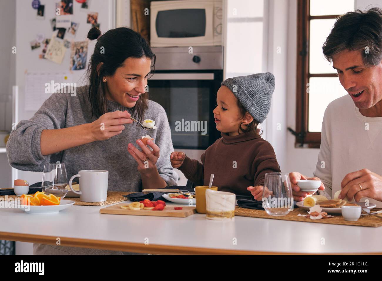 Crop smiling parents with charming girl at table with boiled egg and ...