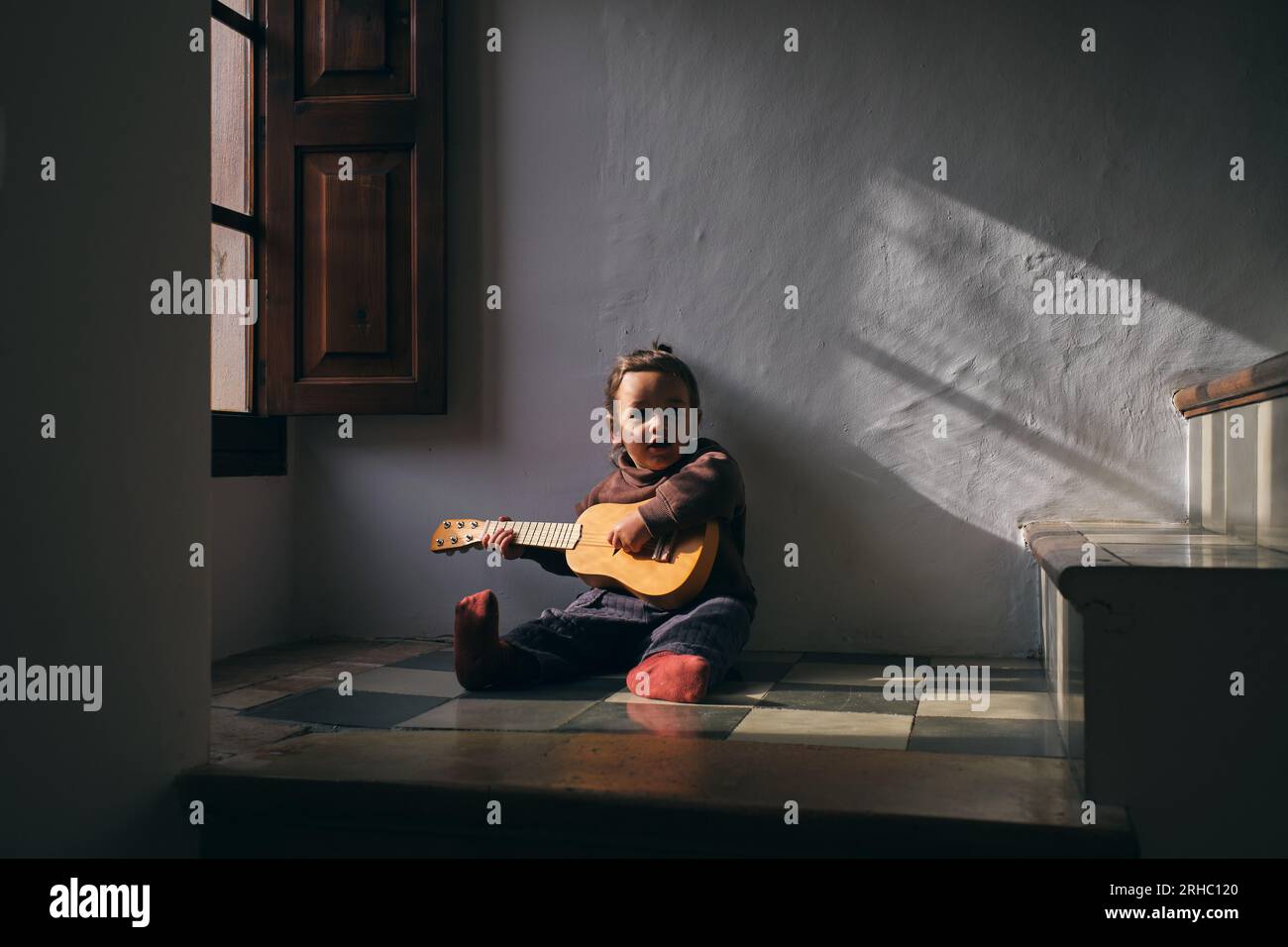 Little musician in casual wear sitting on tiled floor while playing ...