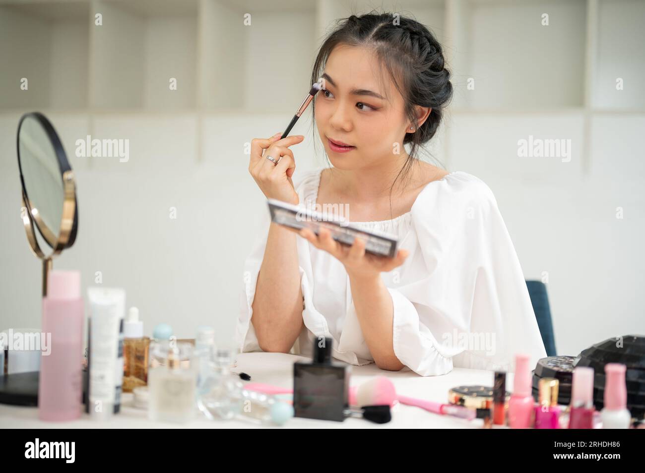 A beautiful and charming Asian woman applies eye shadow to her eyelids, getting ready at her ...