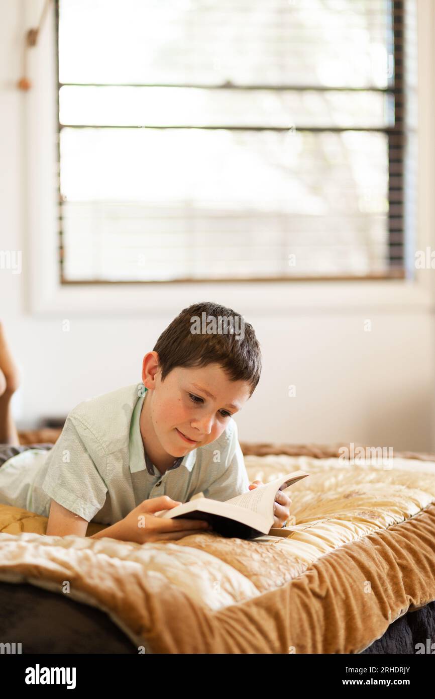 Young boy on bed reading a book with window light coming through blinds ...