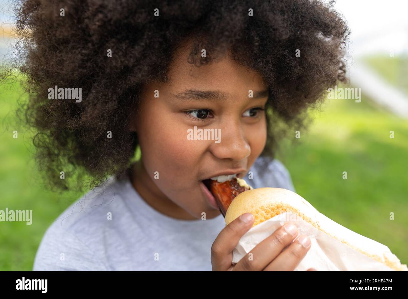 Dark skinned female child eating junk food with pleasure in park while ...