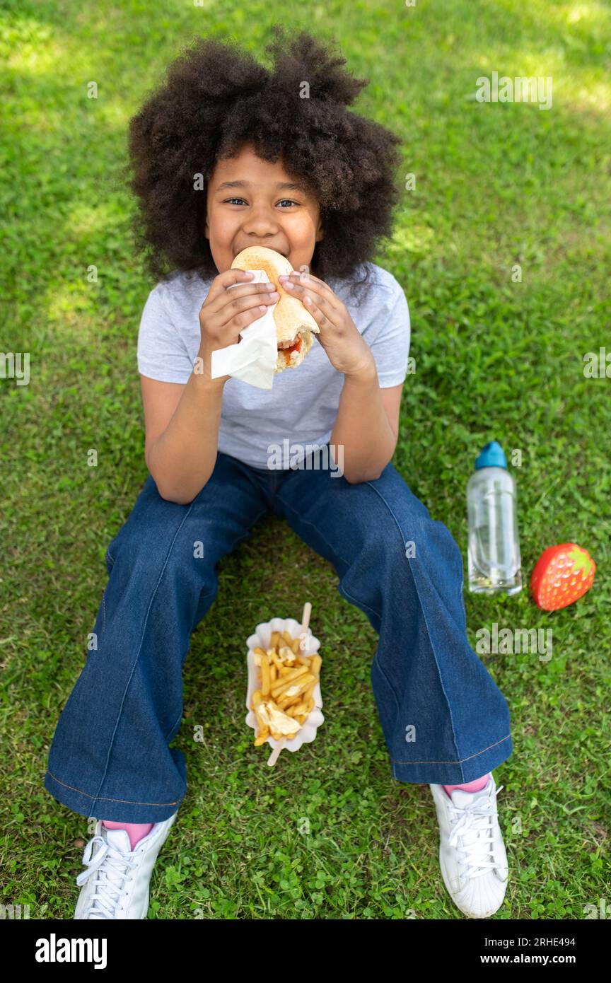 Dark skinned female child eating junk food with pleasure in park while ...