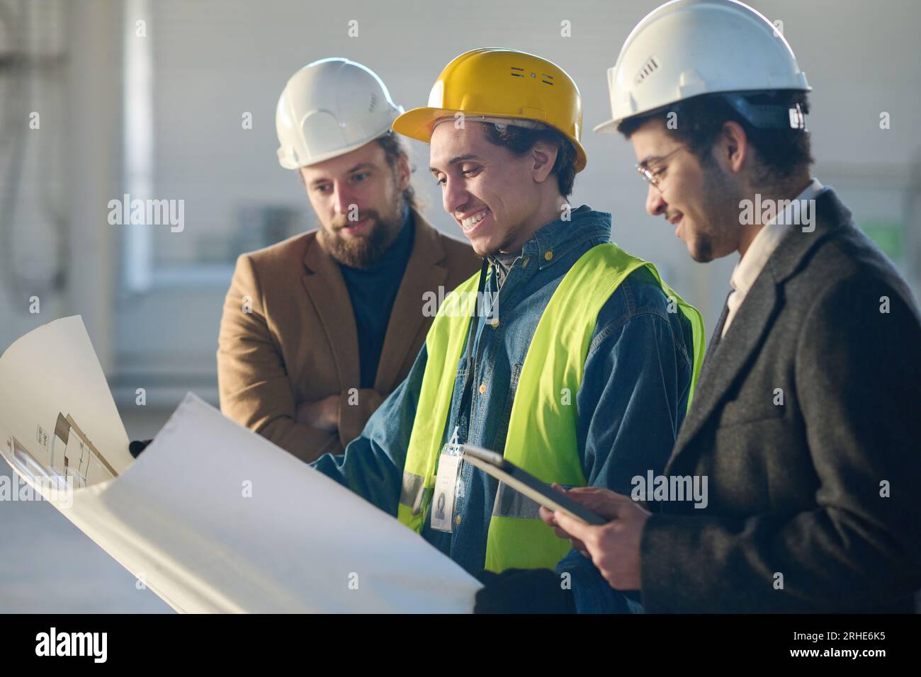 Focus on young smiling engineer in workwear and hardhat holding ...