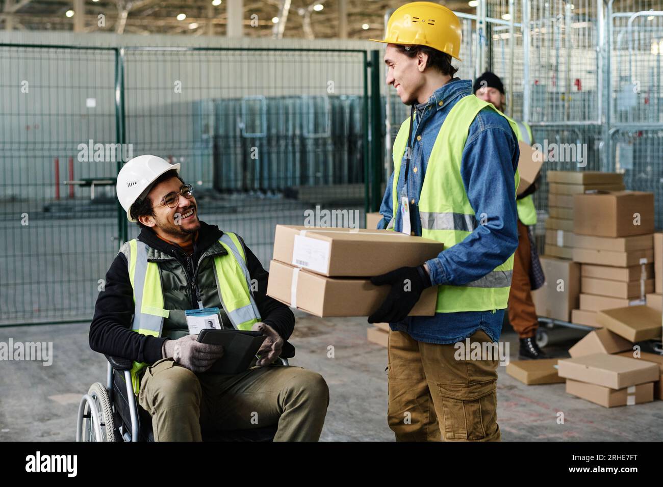 Happy young engineer in wheelchair looking at colleague carrying stack ...