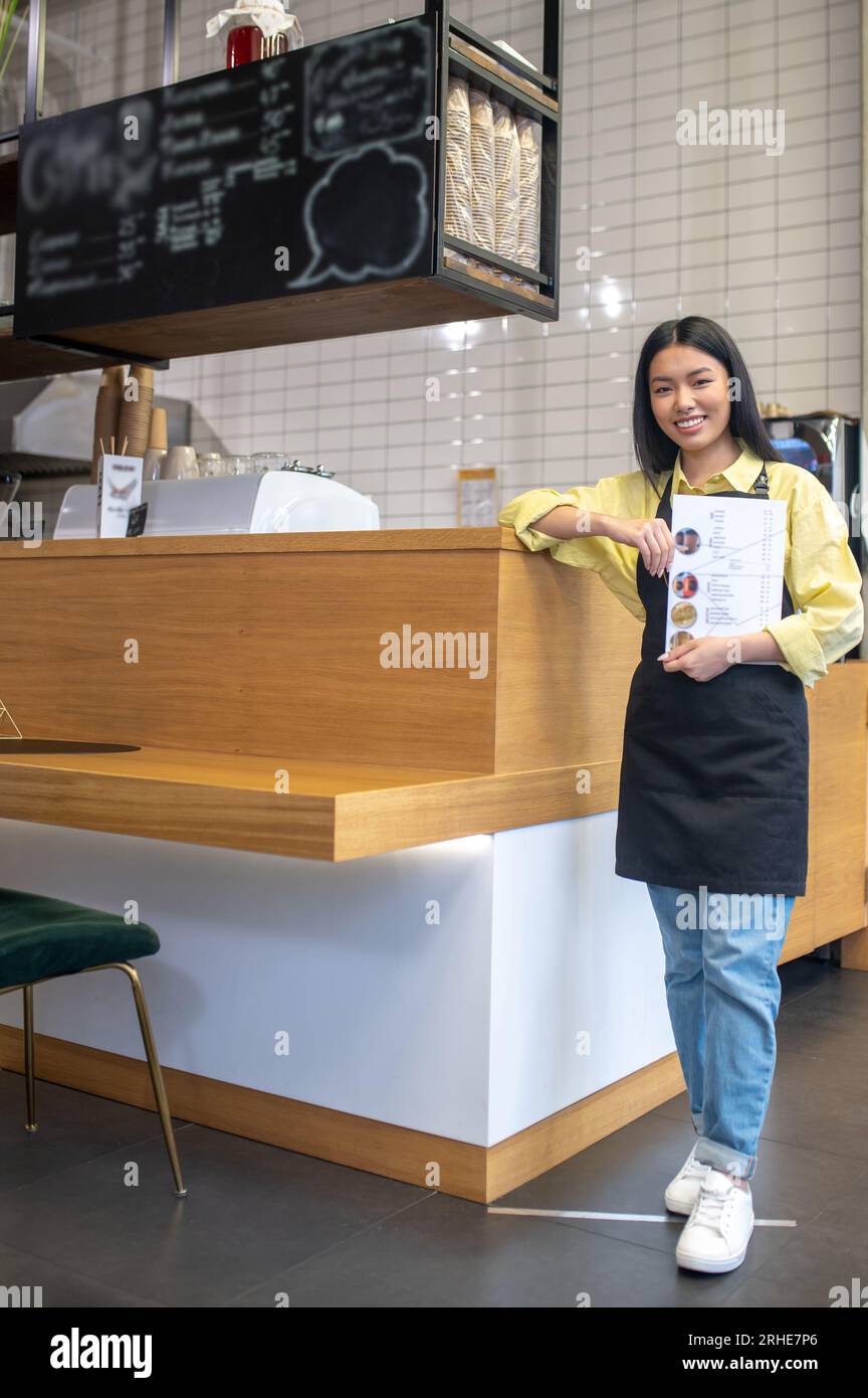 Cute smiling waitress in a black apron standing near the cafe counter ...