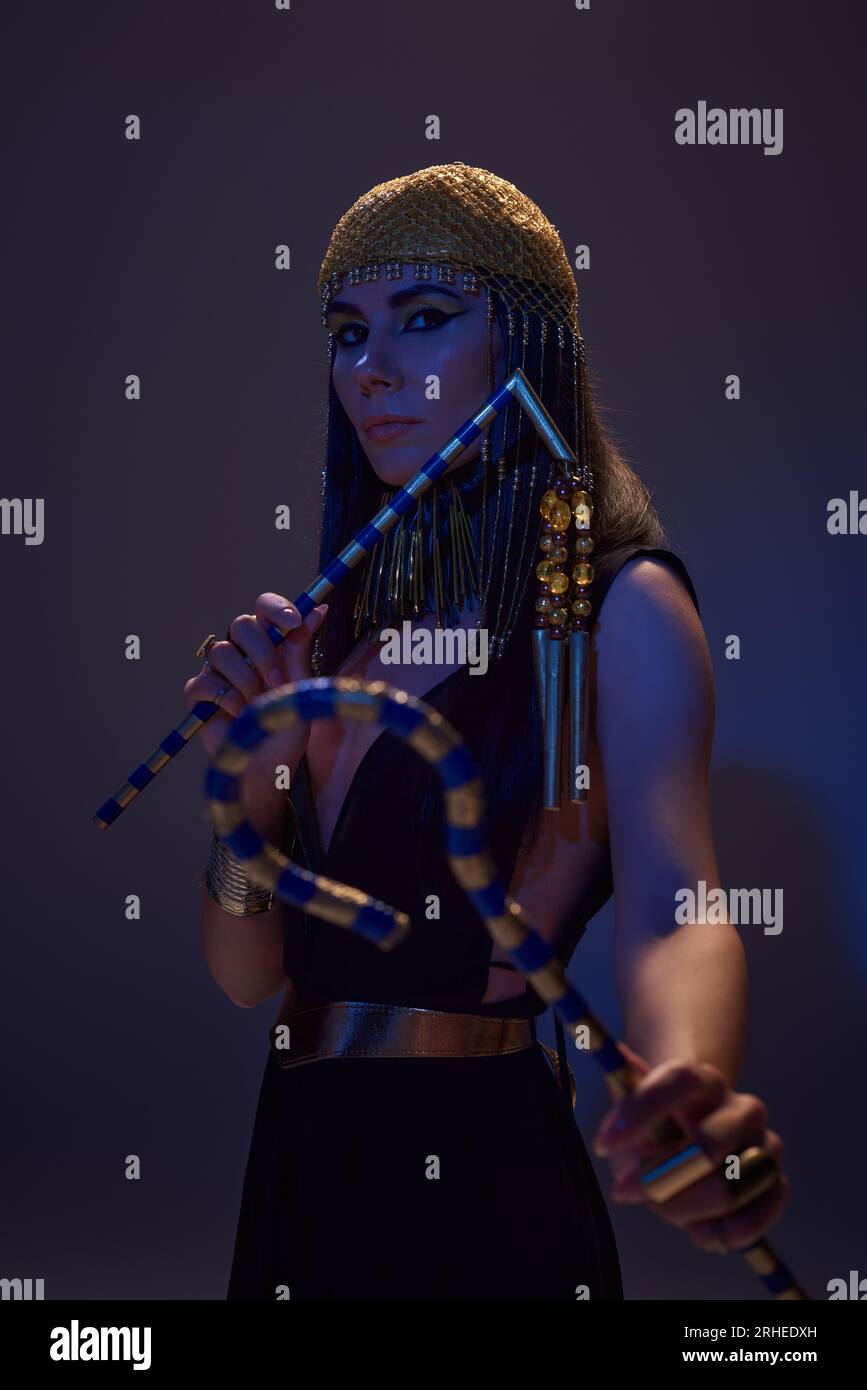 Stylish woman in egyptian attire holding flail and crook in blue light ...