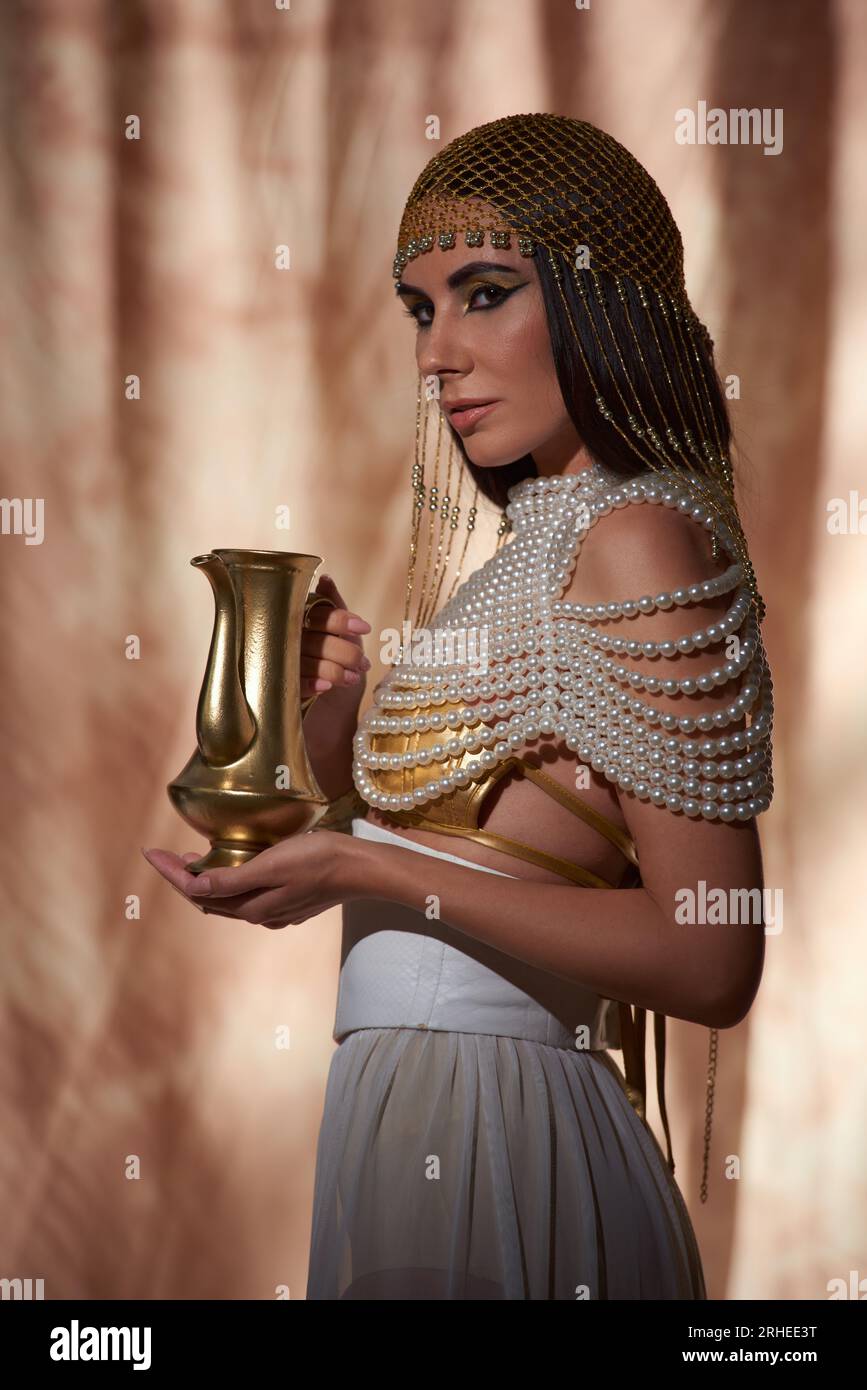 Elegant woman in egyptian headdress and pearl top holding golden jug on ...