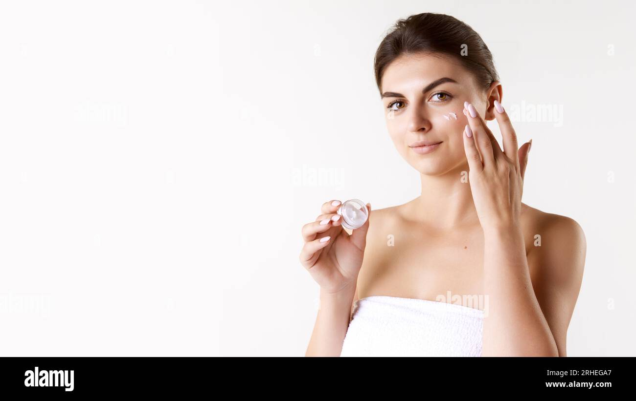 Attractive brunette woman applying under eye cream against white studio ...