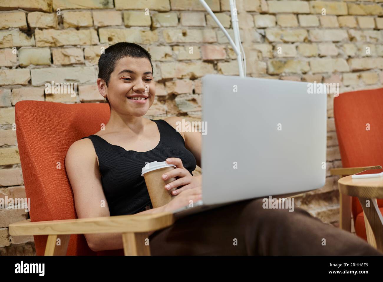 cheerful businesswoman holding coffee to go and using laptop, coworking ...