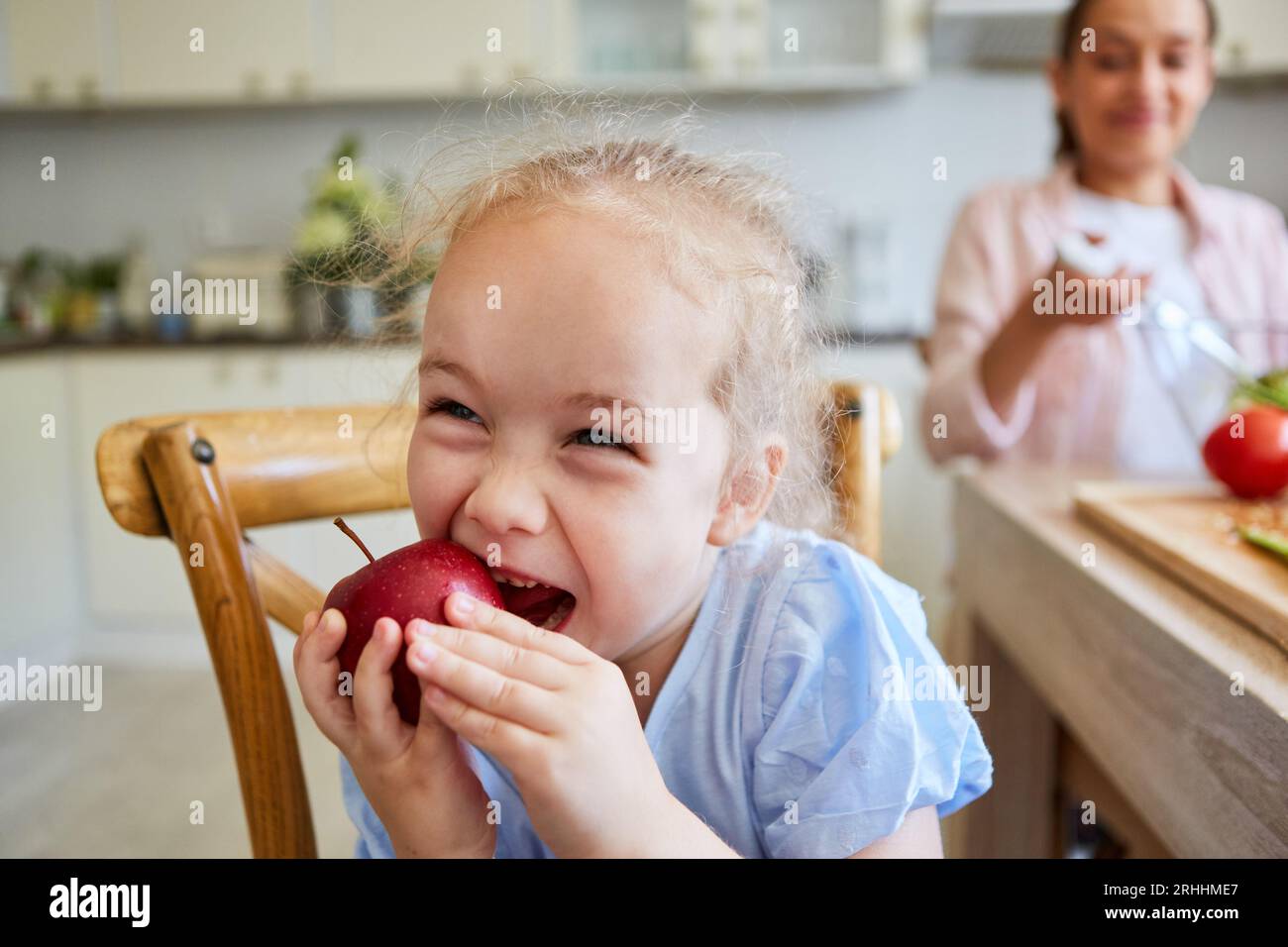 Blond girl biting fresh apple sitting on chair in kitchen at home Stock ...