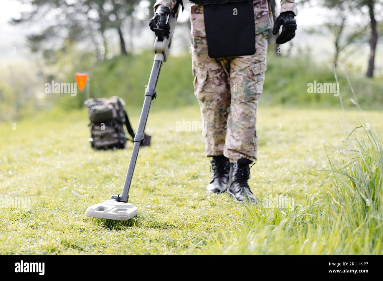 A man in a military uniform and bulletproof vest works in the forest with a metal detector. A ...