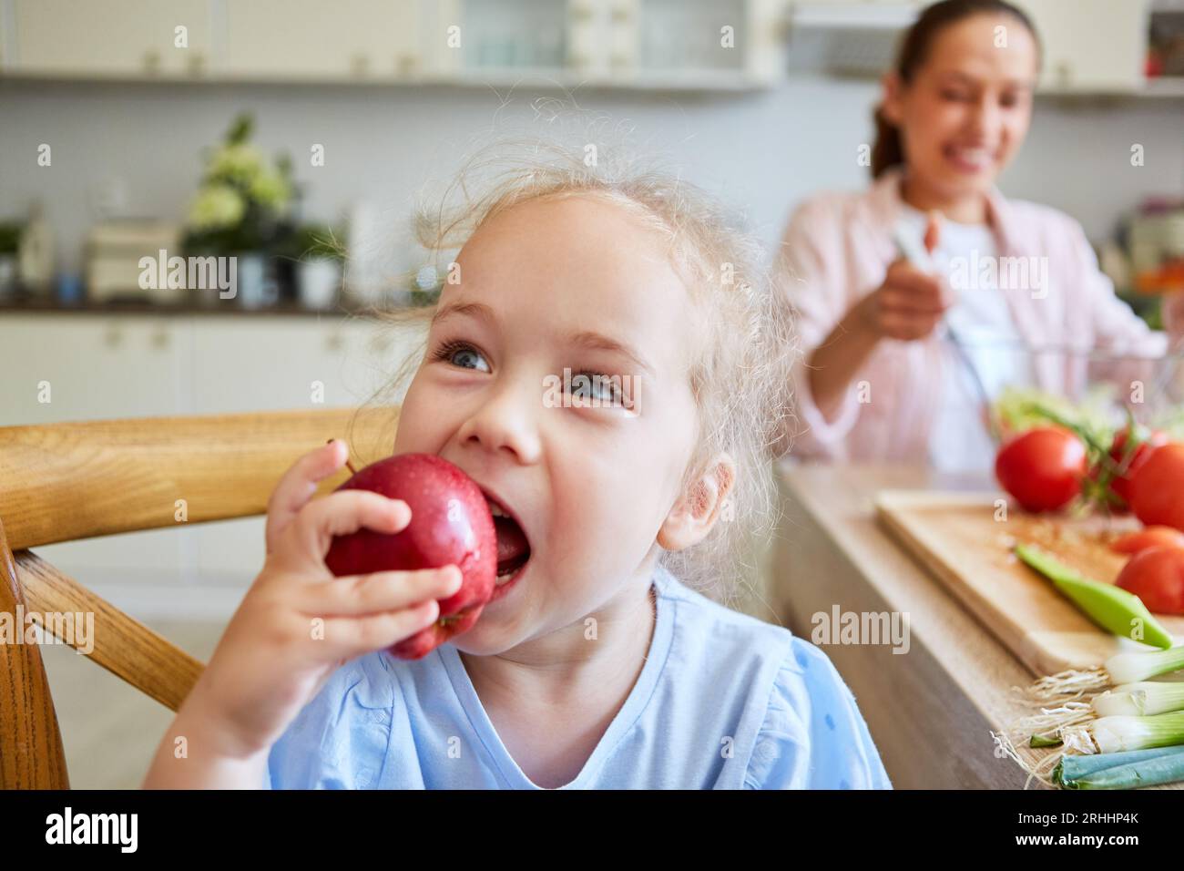 Girl biting in red apple in kitchen with mother watching in the ...