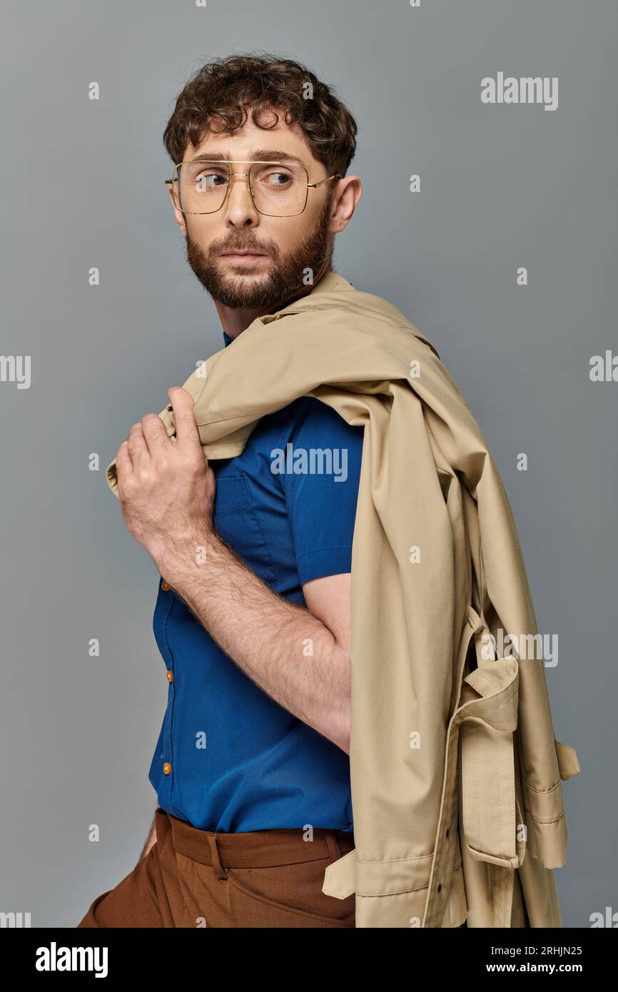 stylish man in glasses holding trench coat, grey backdrop, male model ...