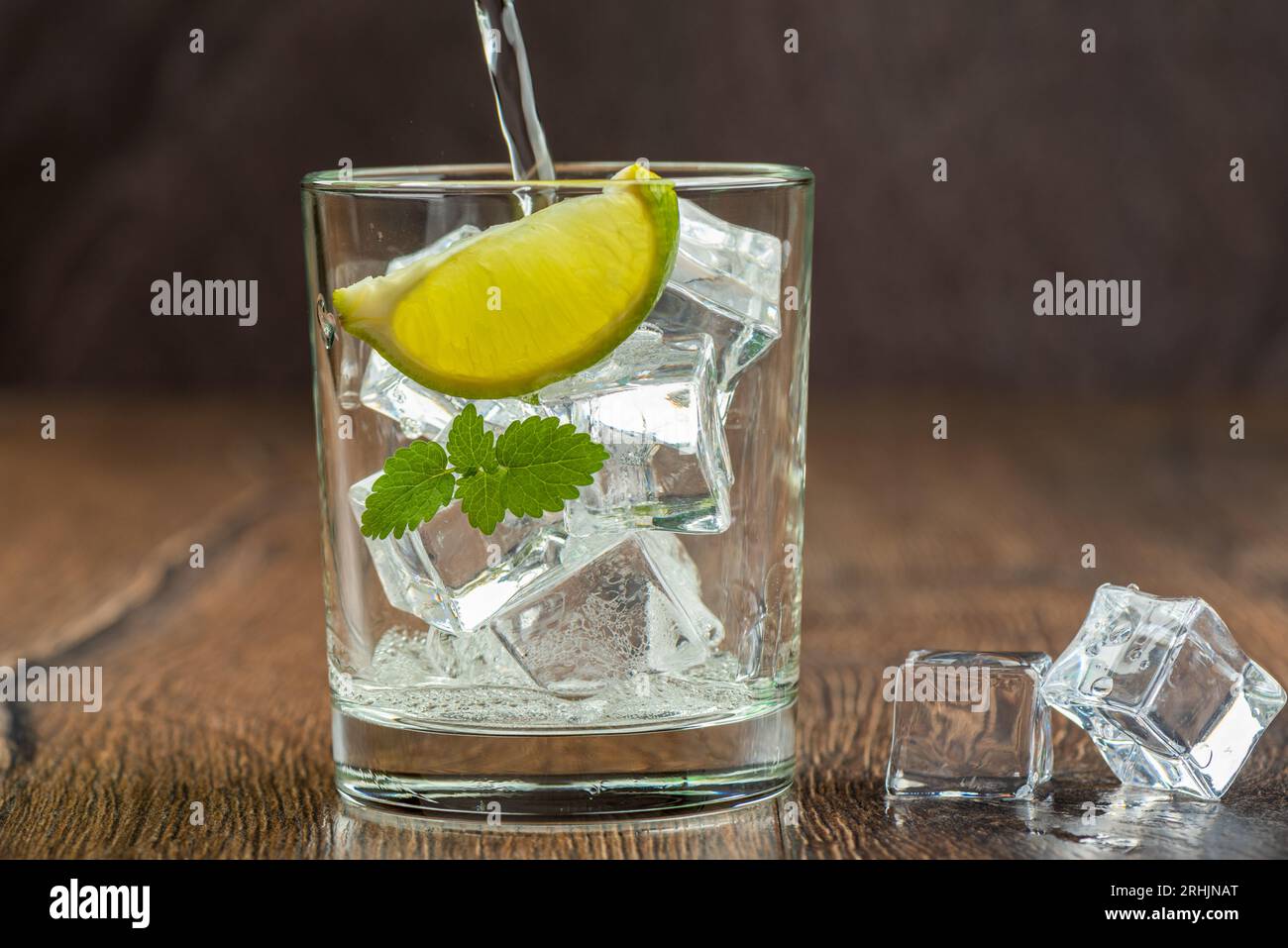 Refreshing water is poured into a glass with ice cubes, lime wedge and mint leaves. Dark wooden ...
