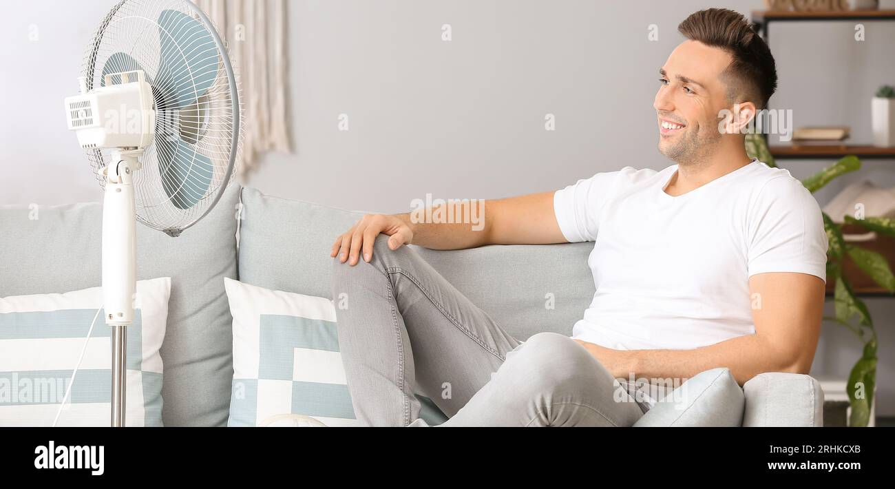 Young man using electric fan during heatwave at home. Banner for design ...