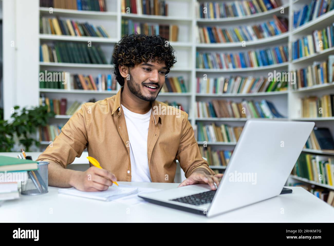 Indian student with curly hair studying sitting among books on shelves ...