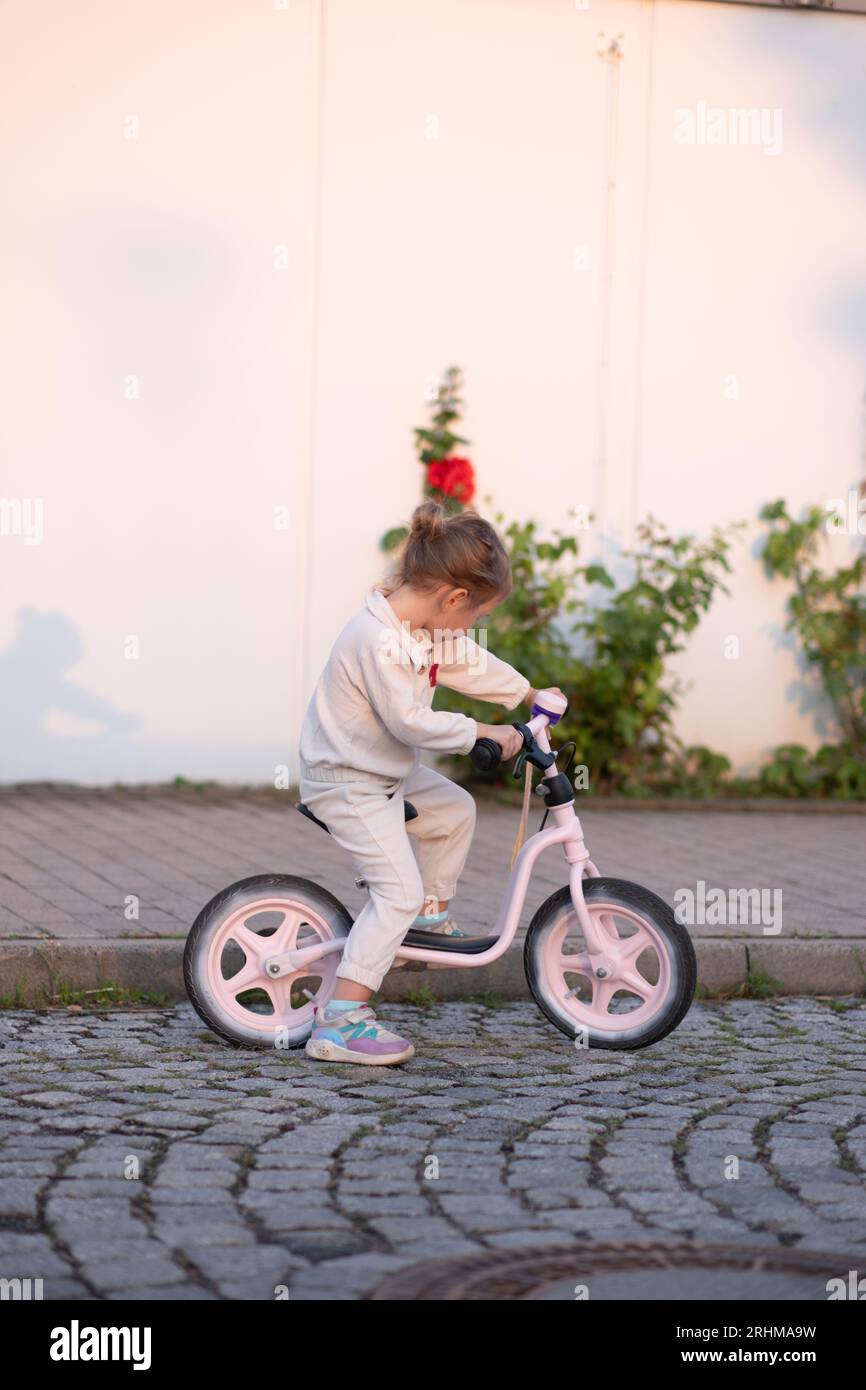 A little girl in a pink suit rides a pink balance bike in the rays of ...