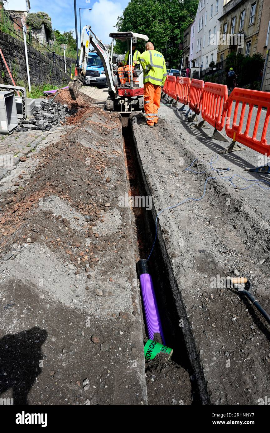 Workmen installing ducting for fibre optic cable in trench cut in road ...