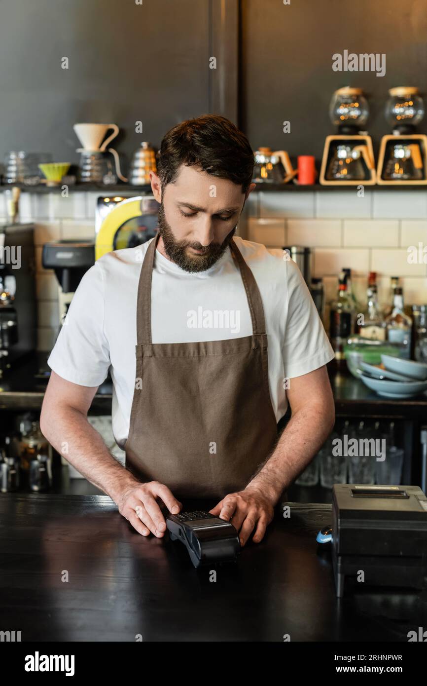 bearded barista in apron using payment terminal on bar while working in ...