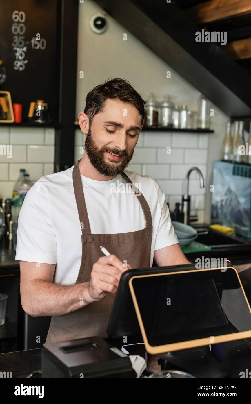 smiling bearded barista in apron using digital tablet while working on ...