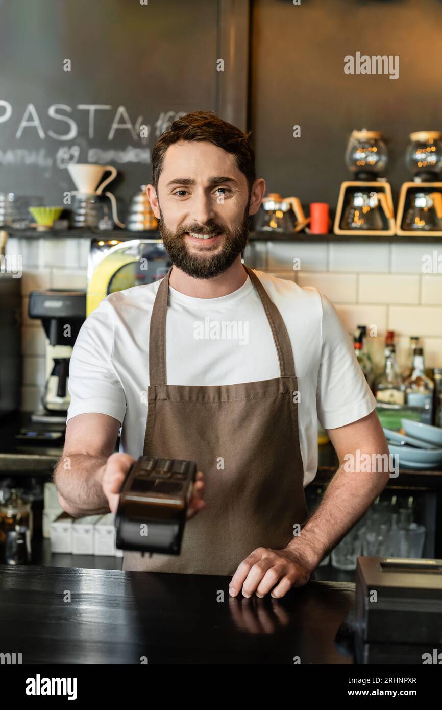 smiling barista in apron holding payment terminal and looking at camera ...