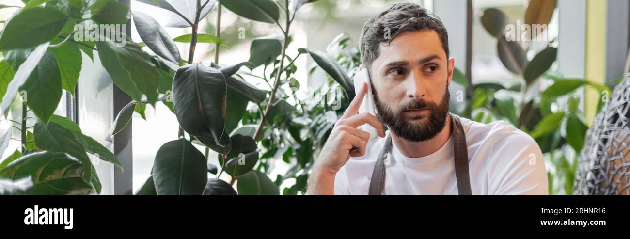 bearded barista in apron talking on smartphone while standing near ...