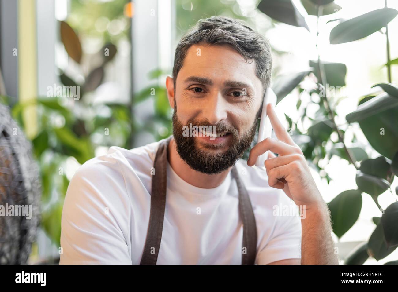 portrait of positive bearded barista talking on smartphone and looking ...