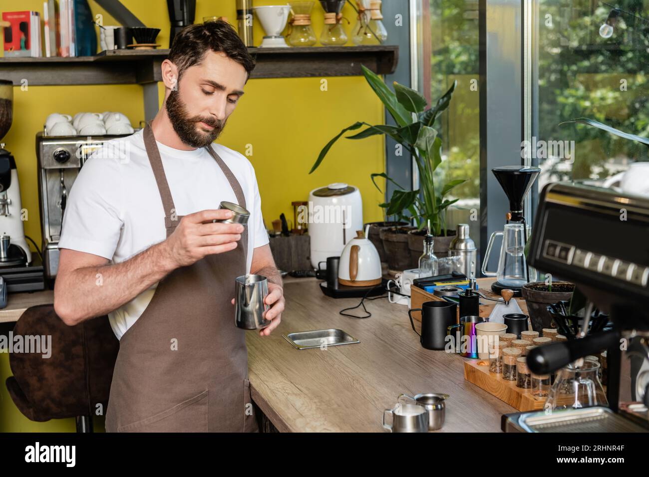 bearded barista in apron holding metal pitchers while making coffee in ...