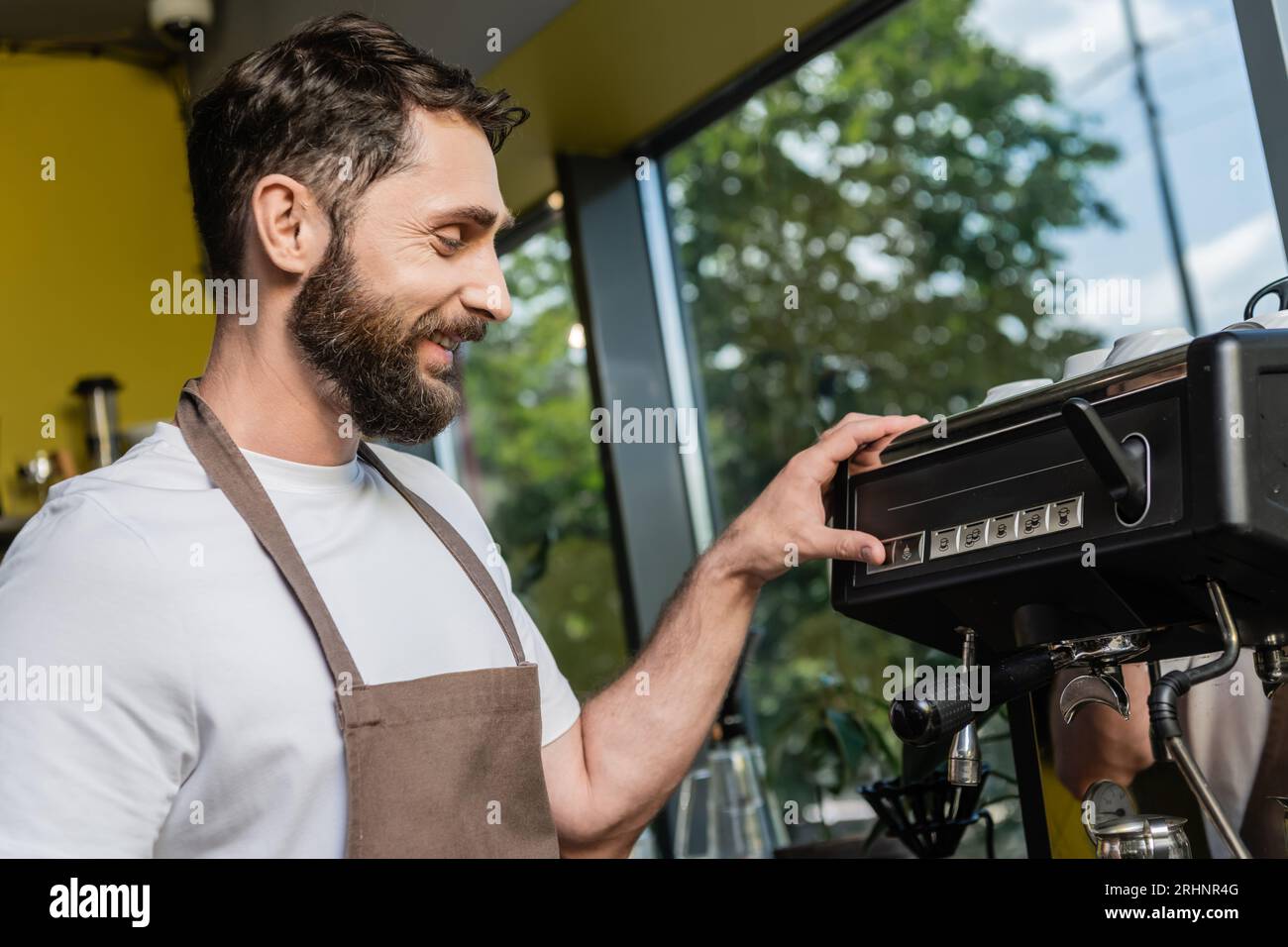 side view of smiling and bearded barista in apron using coffee machine ...