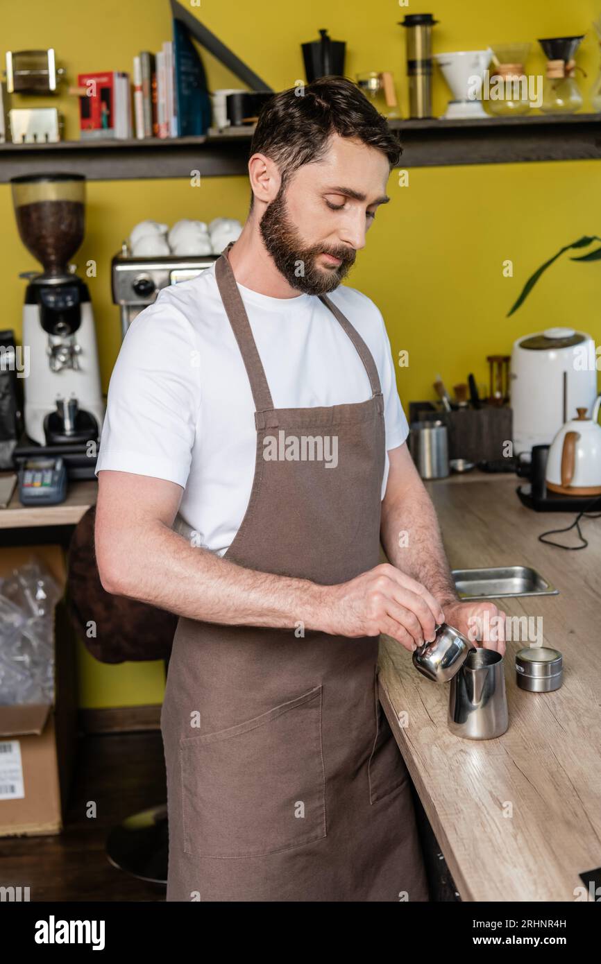 bearded barista in apron pouring coffee in metal pitcher while working ...