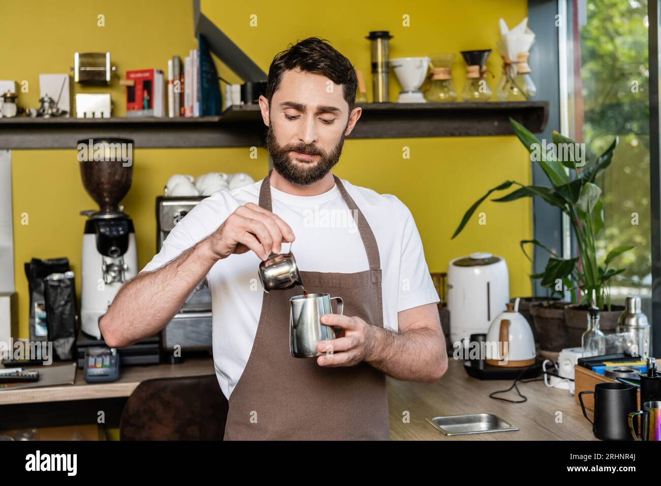 bearded barista in apron pouring coffee in metal pitcher while working ...