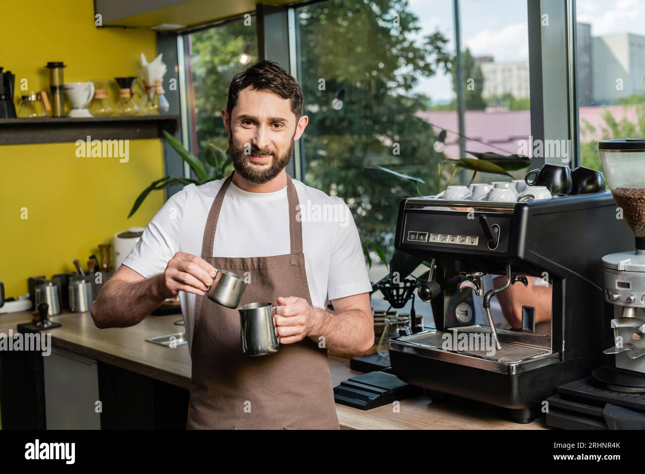 cheerful bearded barista in apron holding pitchers near coffee machine ...