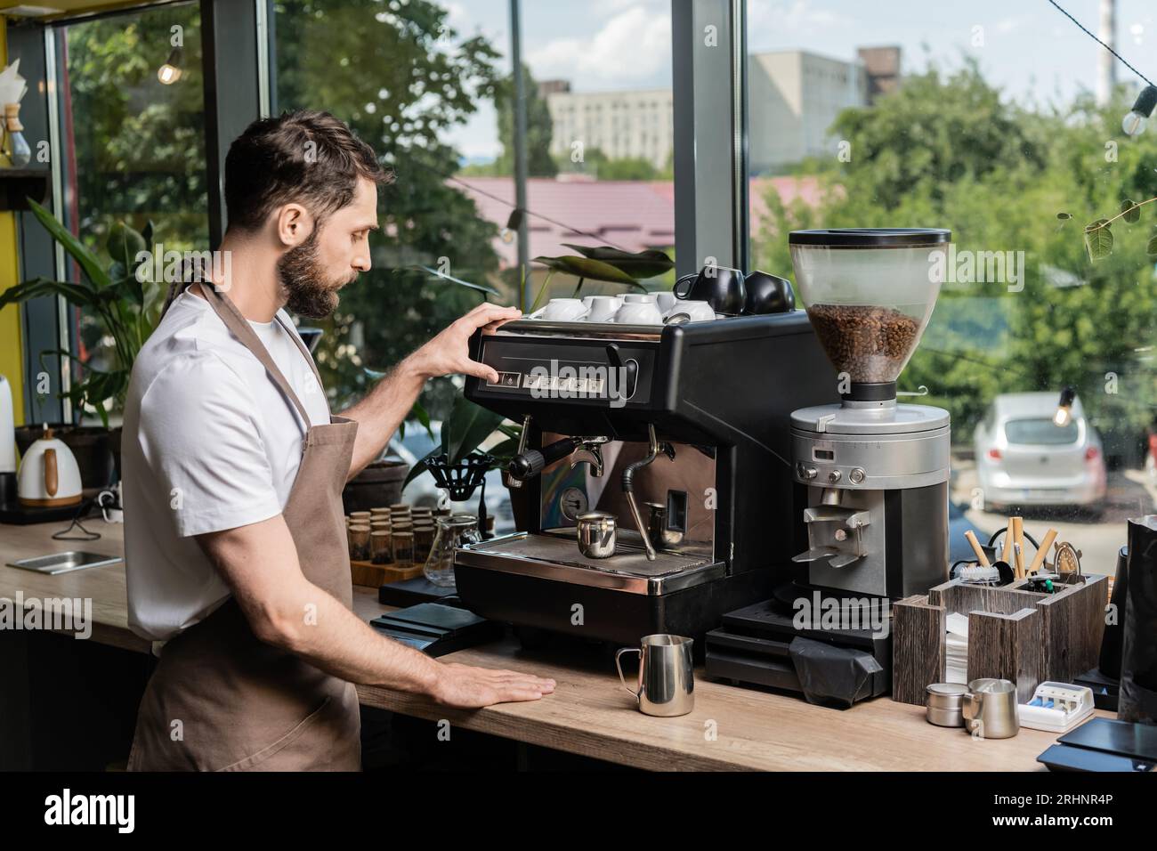 side view of bearded barista in apron making coffee on coffee machine ...