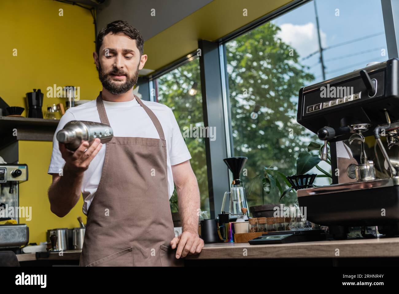 bearded barista in apron using shaker while working near coffee machine ...