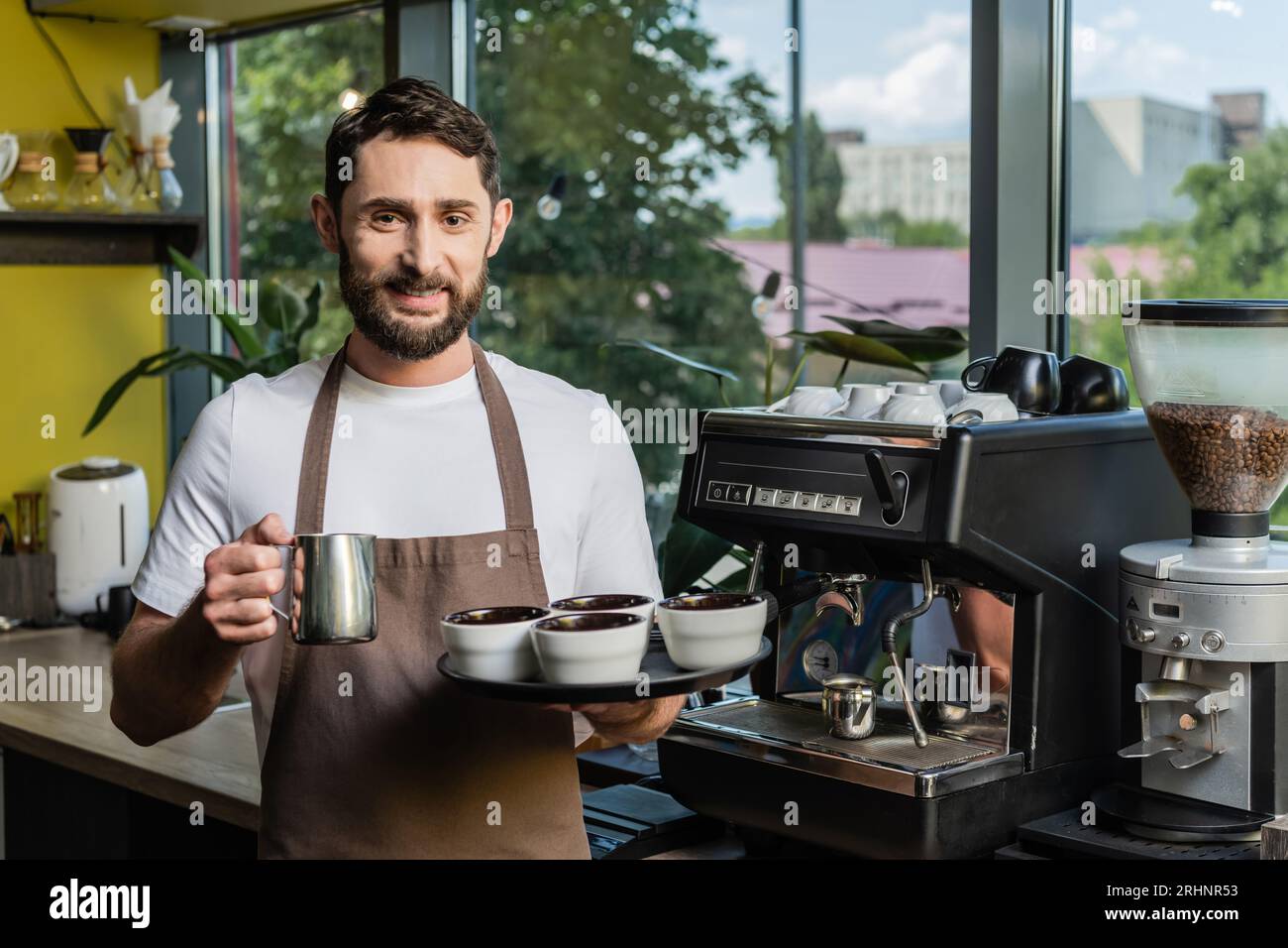 smiling barista in apron holding pitcher and cups near coffee machine ...