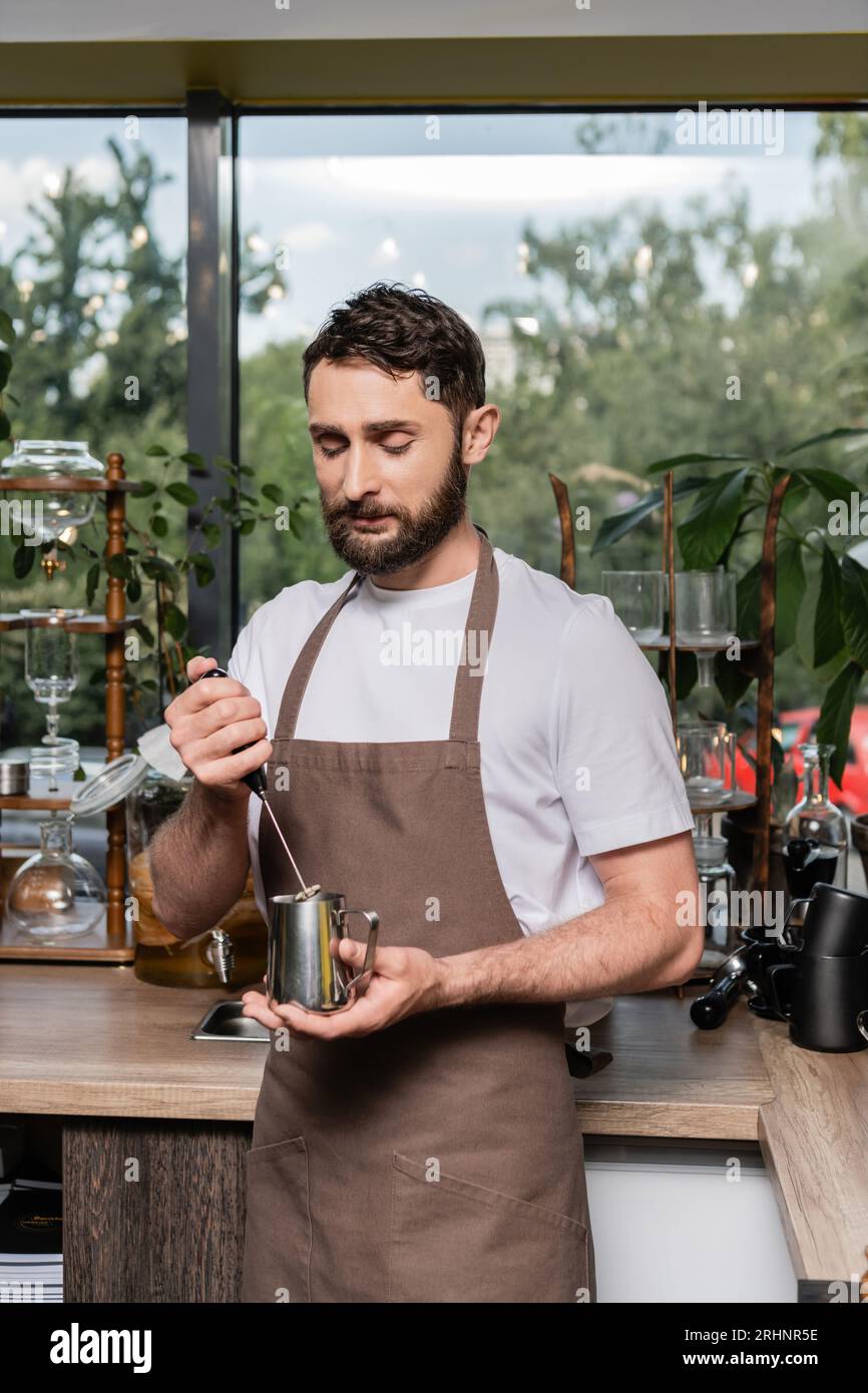 bearded barista in apron holding milk frother and pitcher while working ...