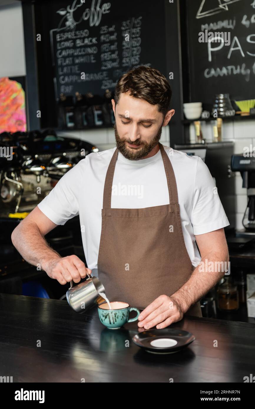 bearded barista in apron pouring milk from pitcher while making ...