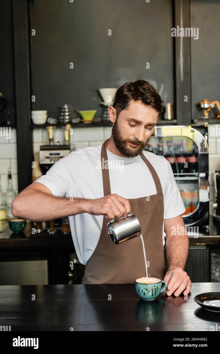bearded barista in apron pouring milk from pitcher while making ...