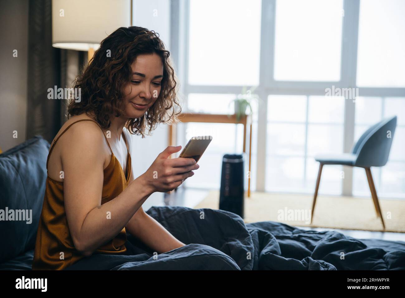 Pretty young woman in her bed with a mobile phone, waking up using a ...