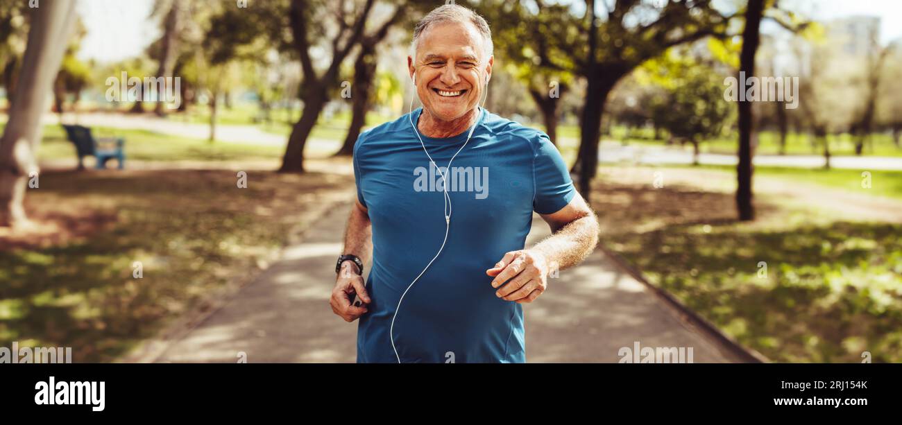 Senior man running in a park with a smile on his face and earphones on ...